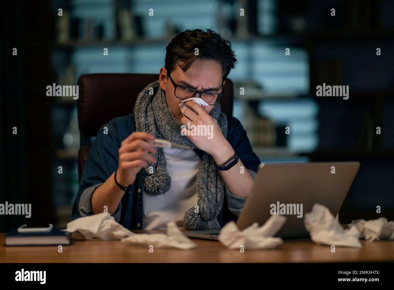 Sick young man sitting in front of computer, holding thermometer Stock ...