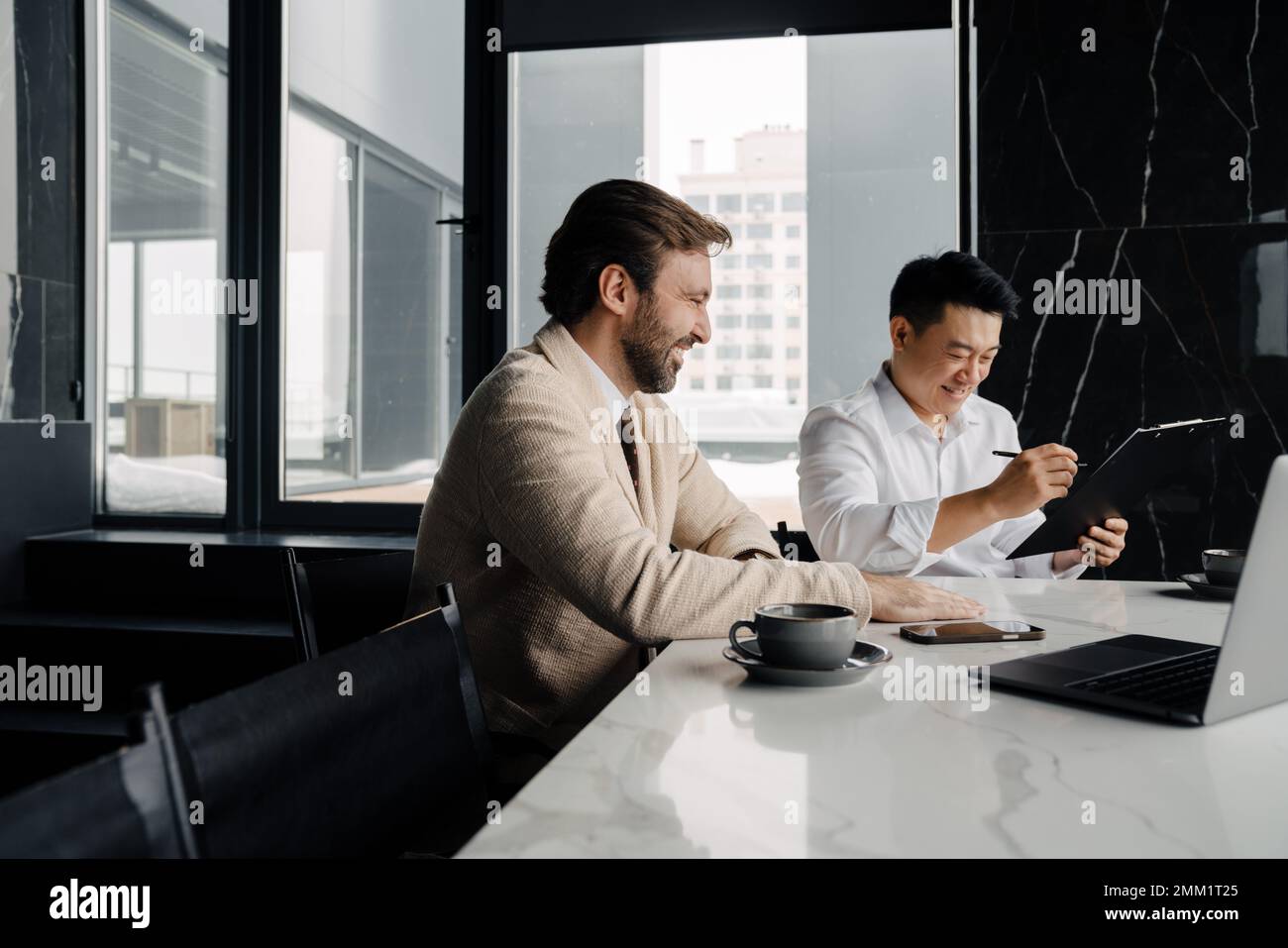Two smiling businessmen sitting by table with laptop one of them ...