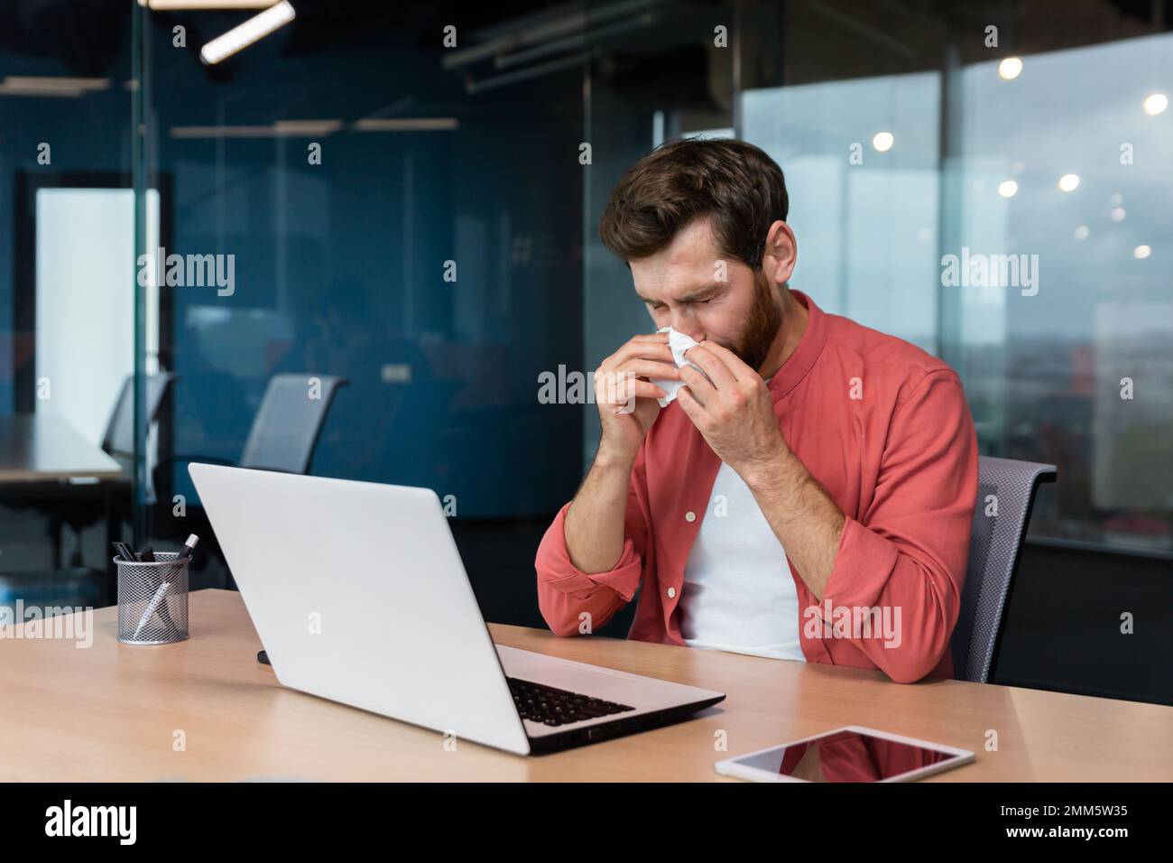 A young man is sick at work. sits at the desk in the office, wipes his ...