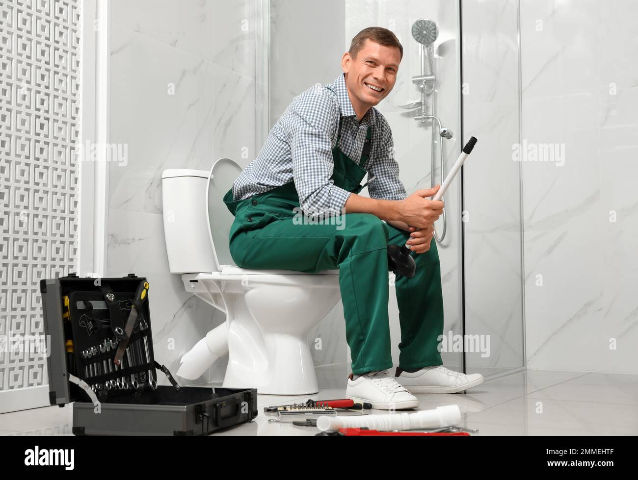 Professional plumber with plunger sitting on toilet bowl in bathroom ...