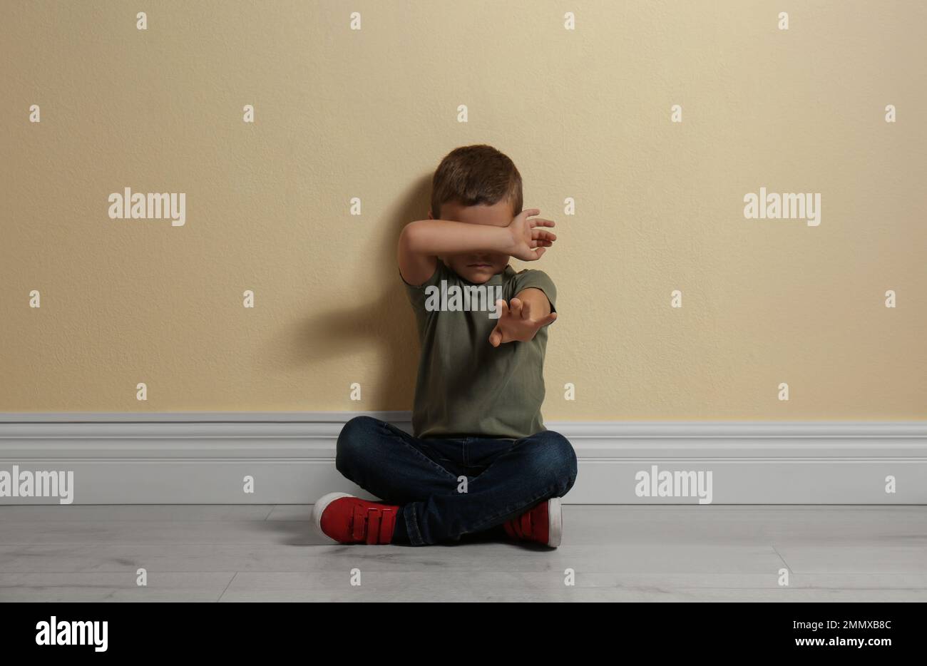 Scared little boy closing face with hand on floor near yellow wall ...