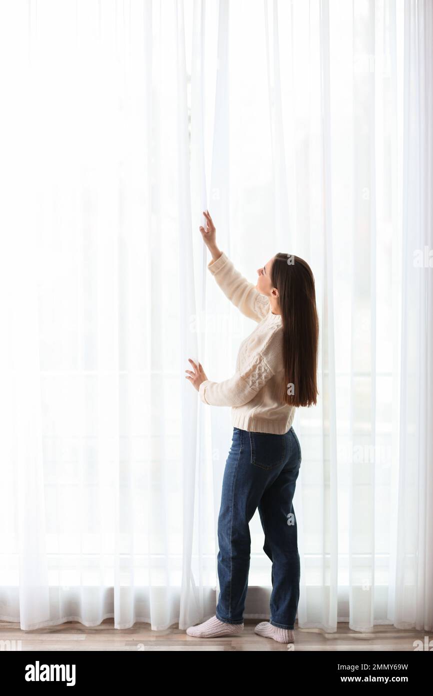 Young woman opening window curtains at home Stock Photo - Alamy