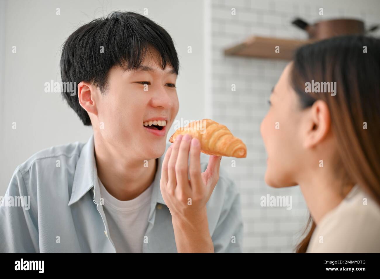 Happy and playful young Asian boyfriend feeding a croissant to his ...