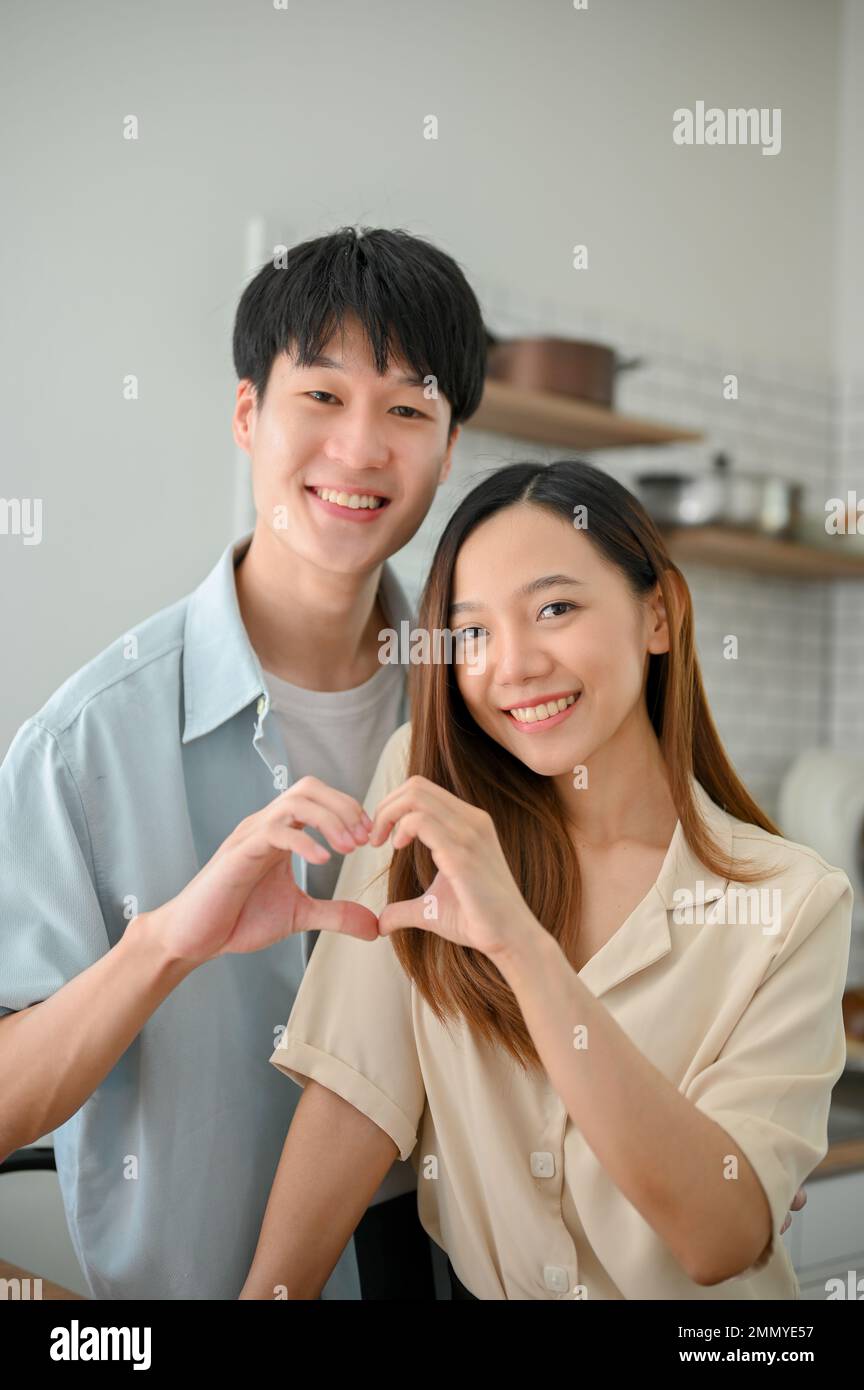 Portrait, Lovely young Asian couple making a heart hand sign together in the kitchen, smiling ...