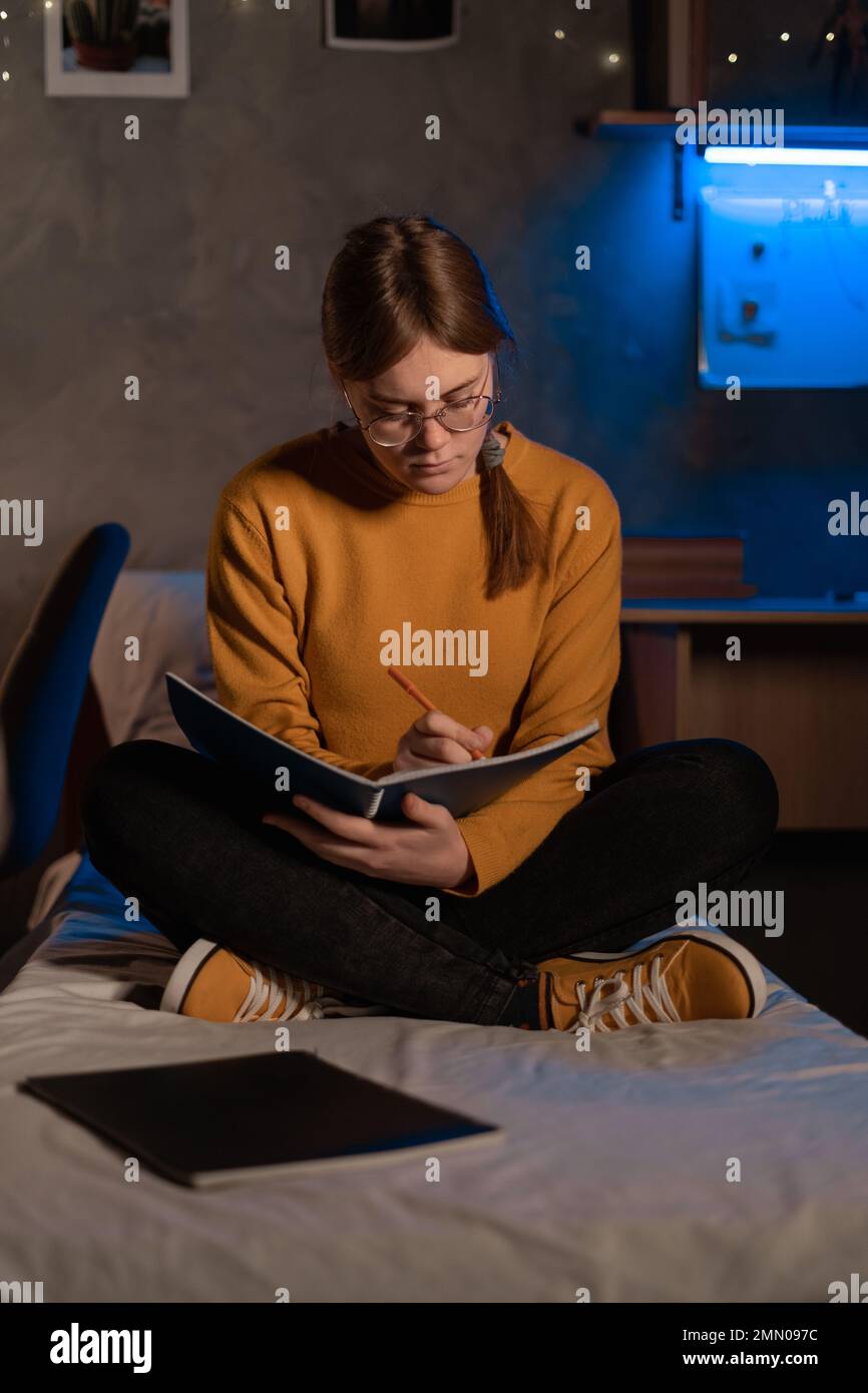 Female university student sitting on bed doing homework using copybooks ...
