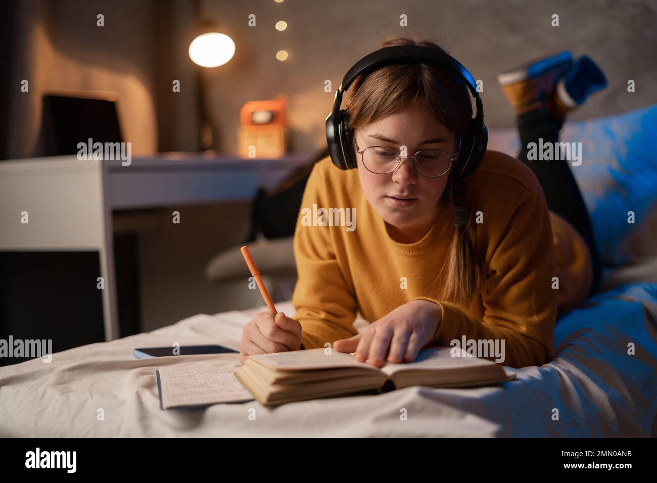Serious student girl in glasses reading book on bed in dormitory ...