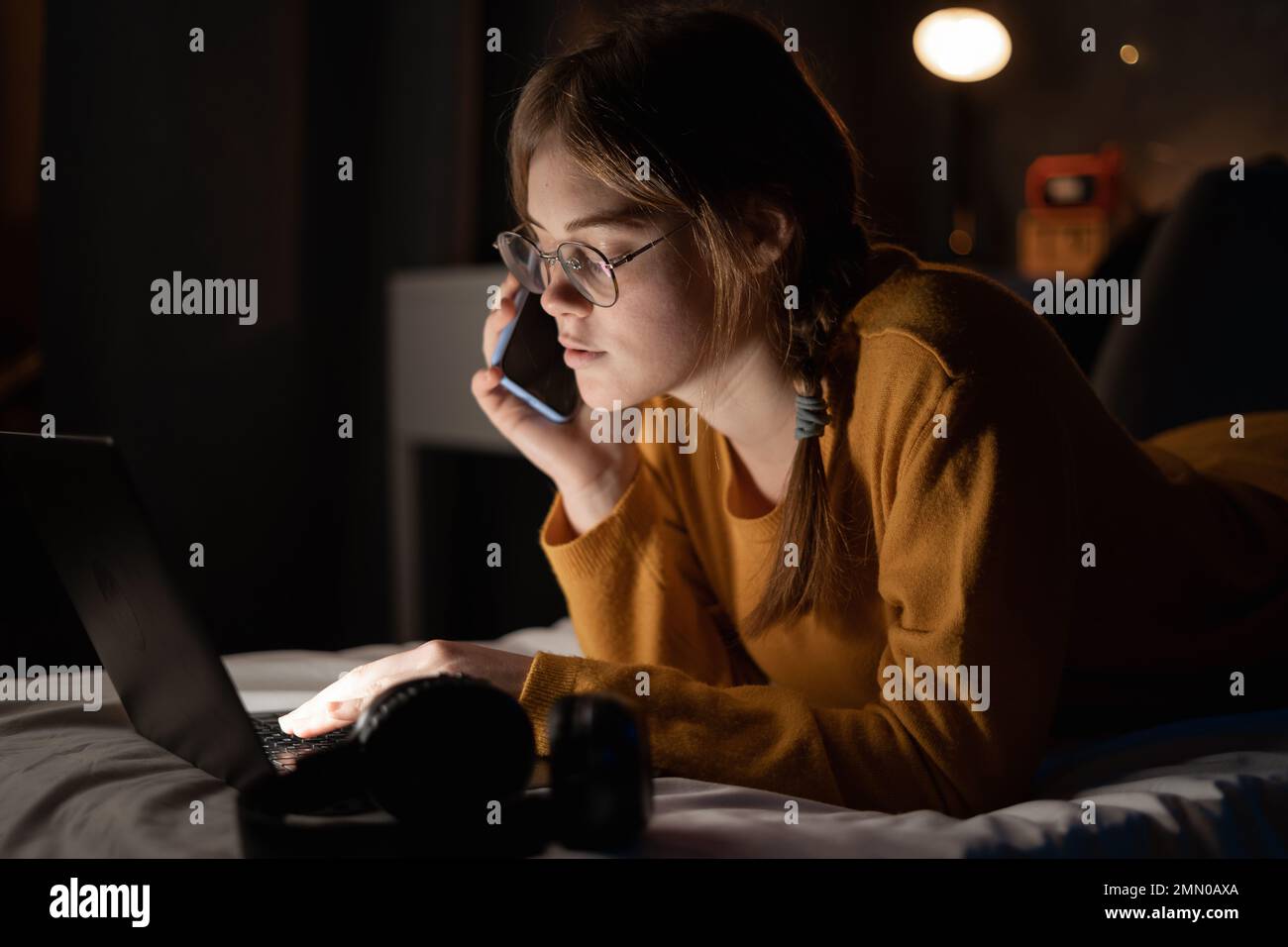 Female college student calling her friends, lying on bed working on ...