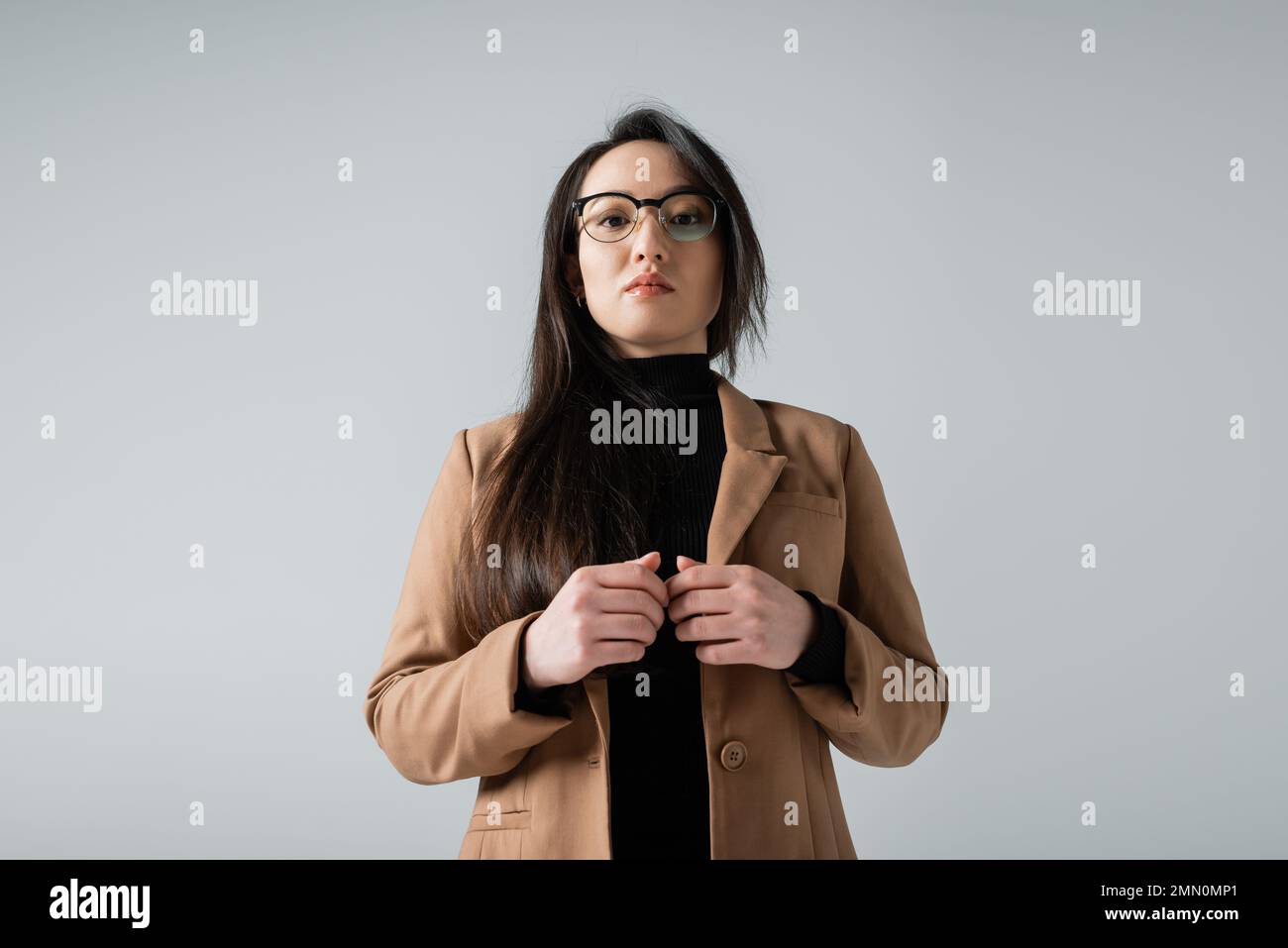 young asian woman in beige blazer and black turtleneck looking at camera isolated on grey Stock ...