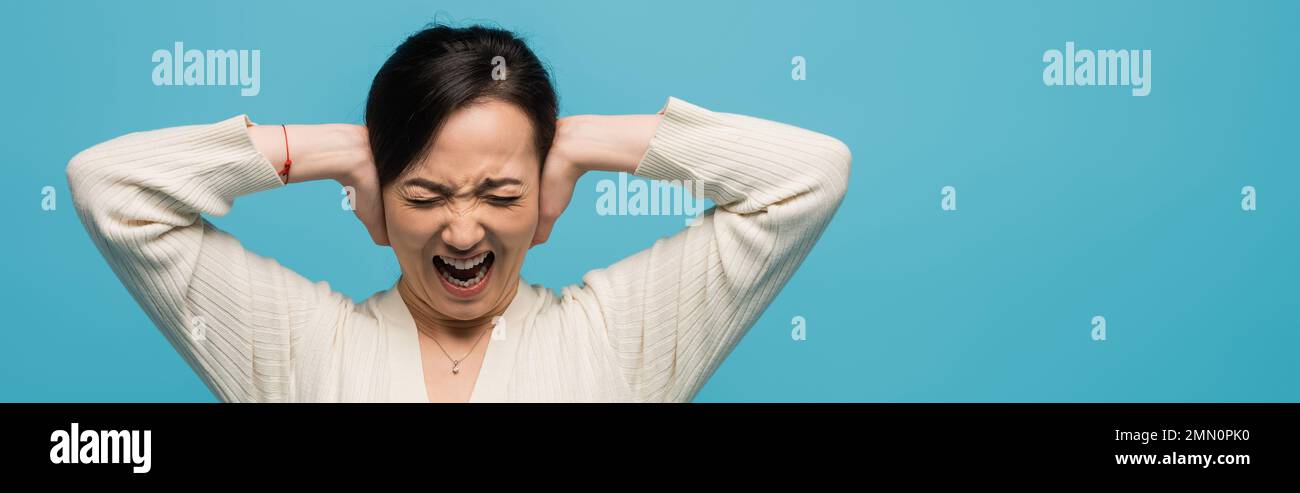 Aggressive asian woman screaming and covering ears isolated on blue ...