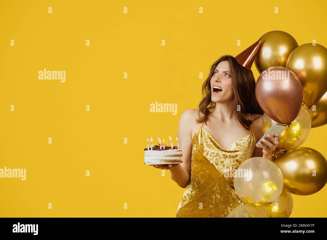 Surprised woman with balloons, yummy cake and smartphone, looking aside ...