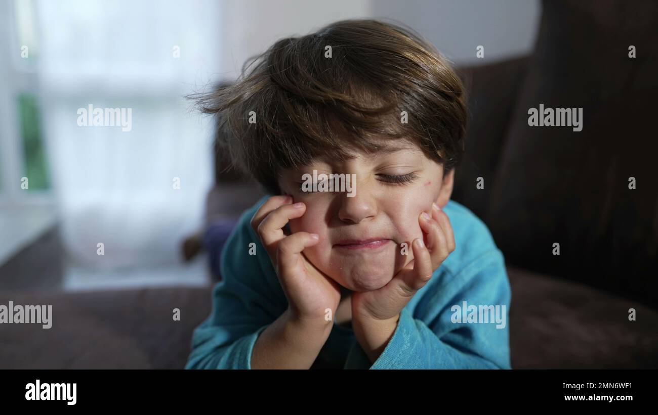 One upset small boy grimacing with hand in chin laying on couch at home ...