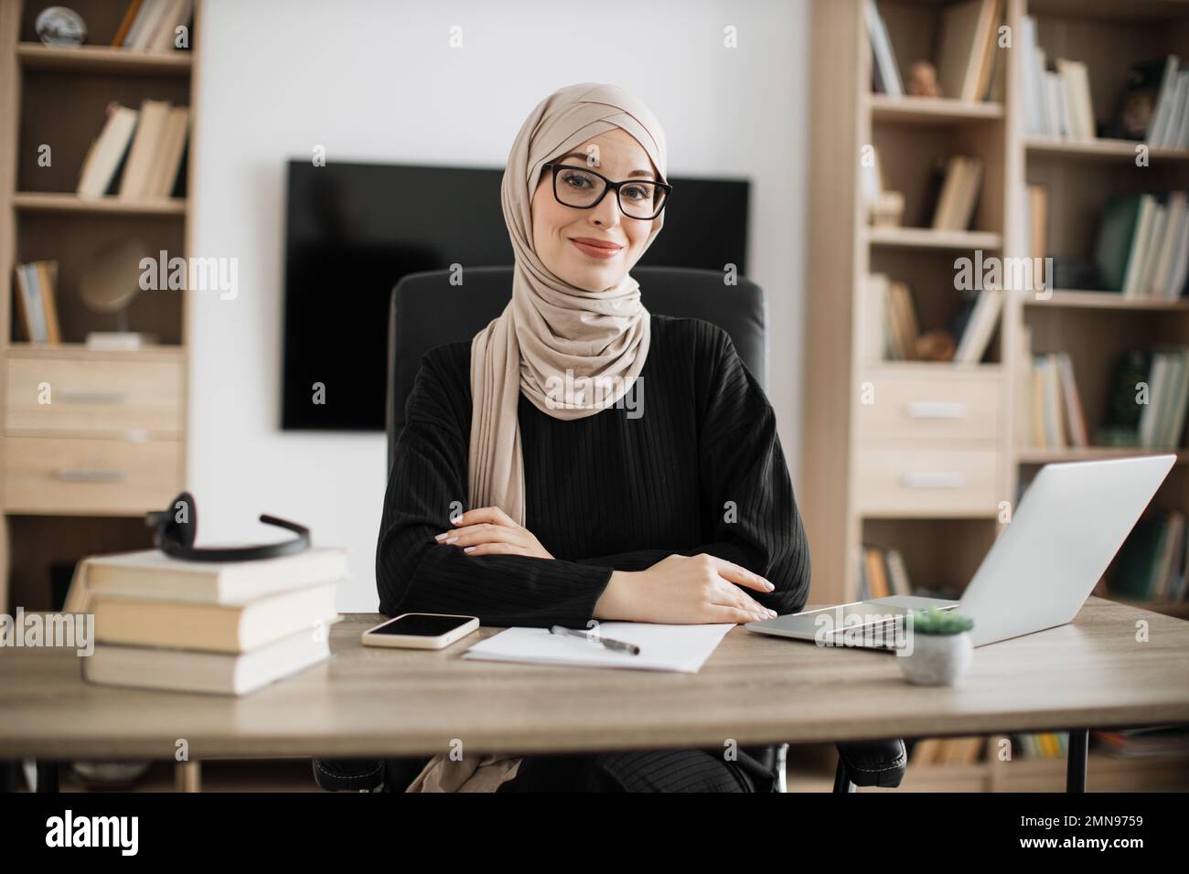 Smiling muslim woman in eyeglasses and hijab sitting at desk with books ...