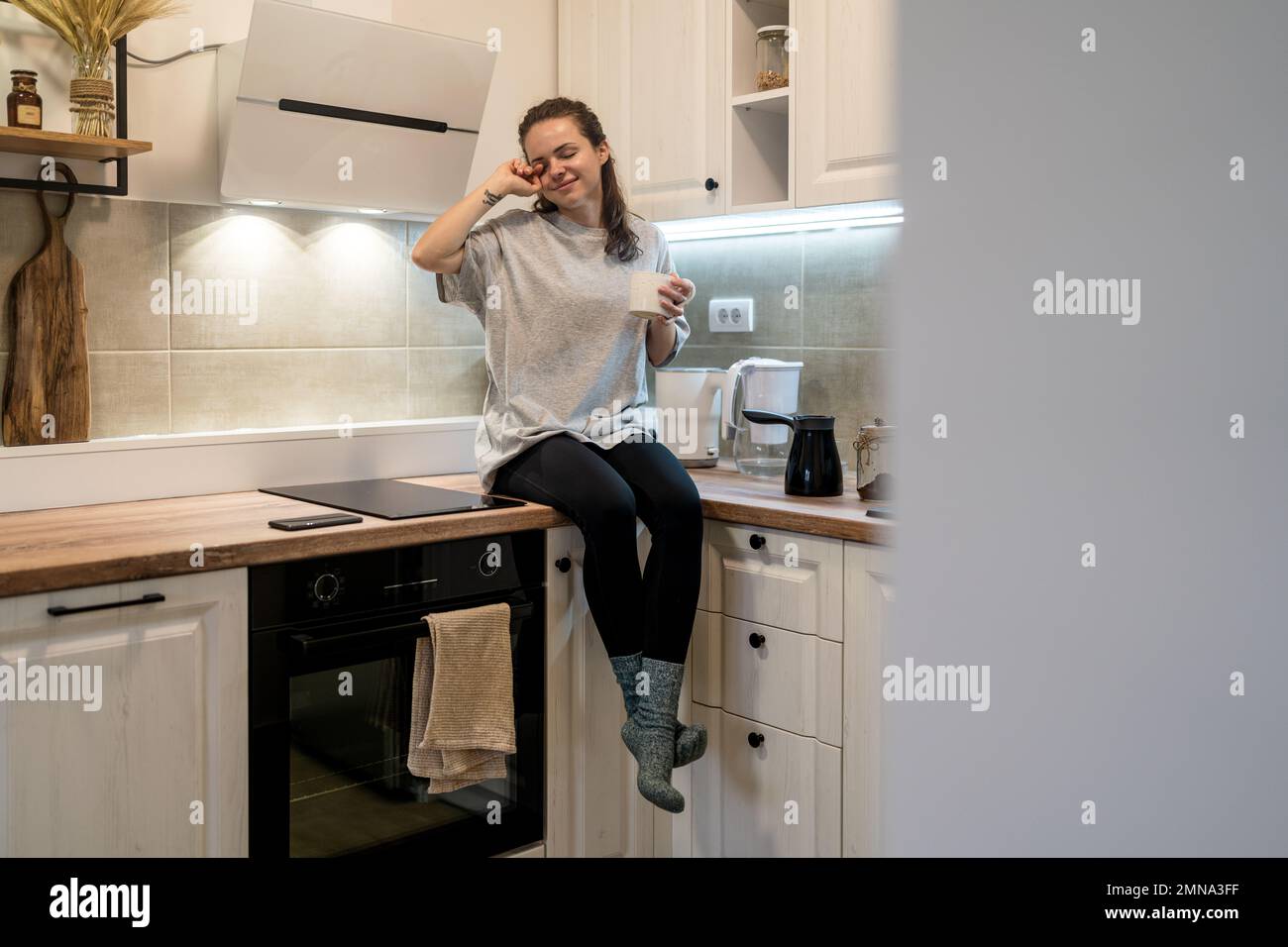 Woman sitting in kitchen in the morning holding mug of coffee rubbing ...