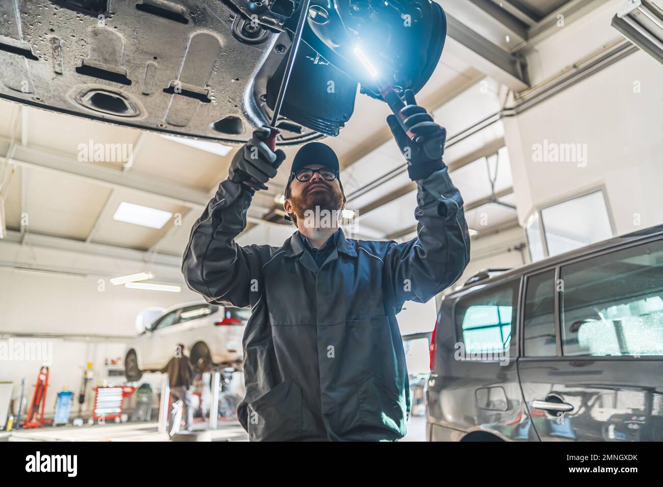 An expert mechanic with an LED light inspects a car on a lift in a ...