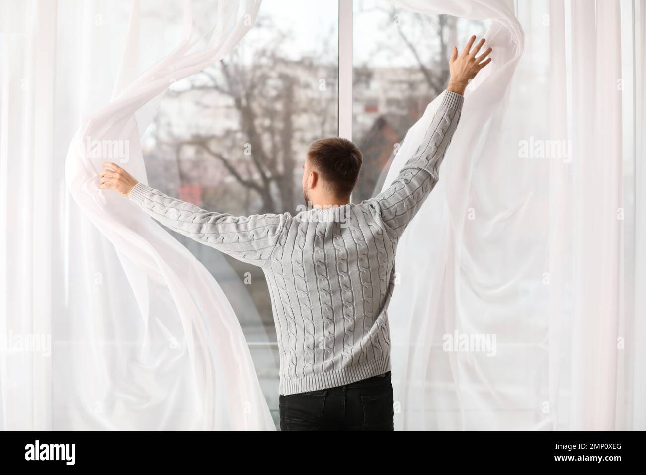Young man opening window curtains at home, back view Stock Photo - Alamy