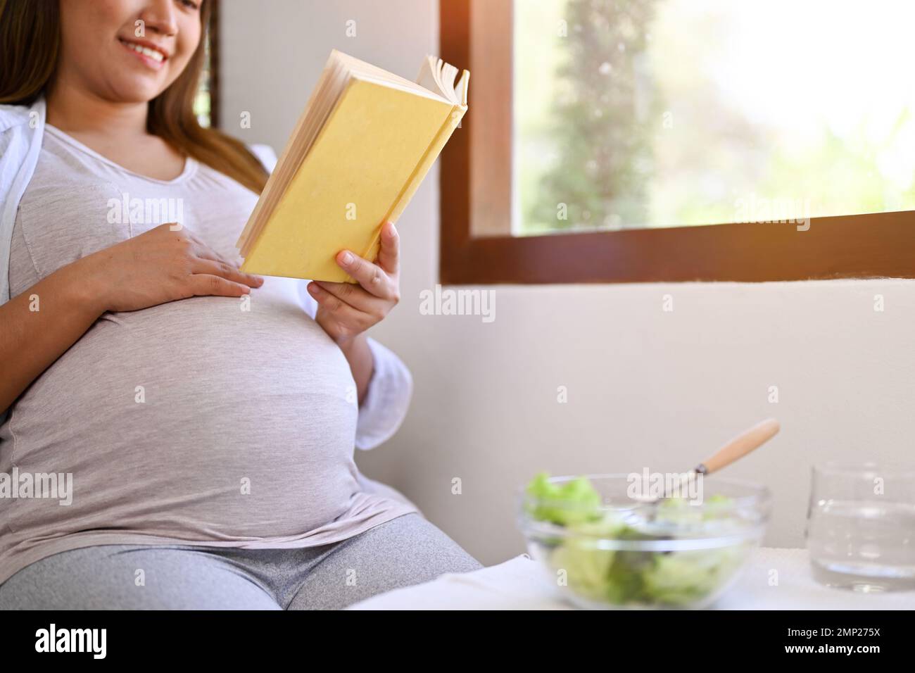 Happy and relaxed Asian pregnant woman touching her belly, reading a ...