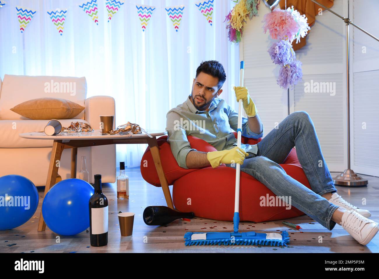 Young man with mop suffering from hangover in messy room after party ...