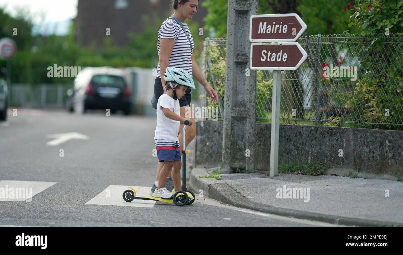 mother and child crossing street on sidewalk. Kid wearing helmet riding ...