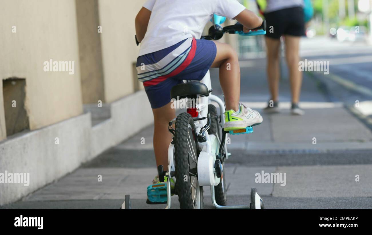 Back of child rides bike with supportive wheels outside in city street ...