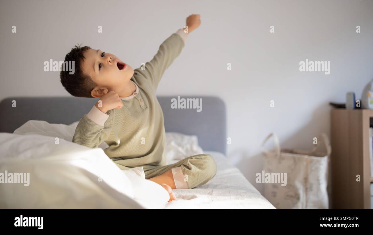Cheerful active caucasian little child wake up, sits on bed, stretching ...