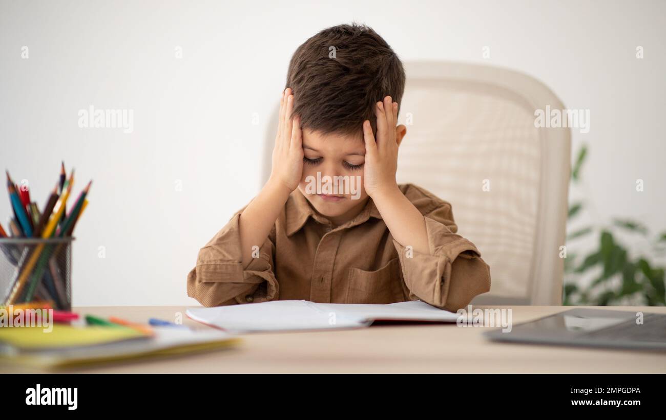 Unhappy sad tired caucasian little child sits at table presses hands to ...