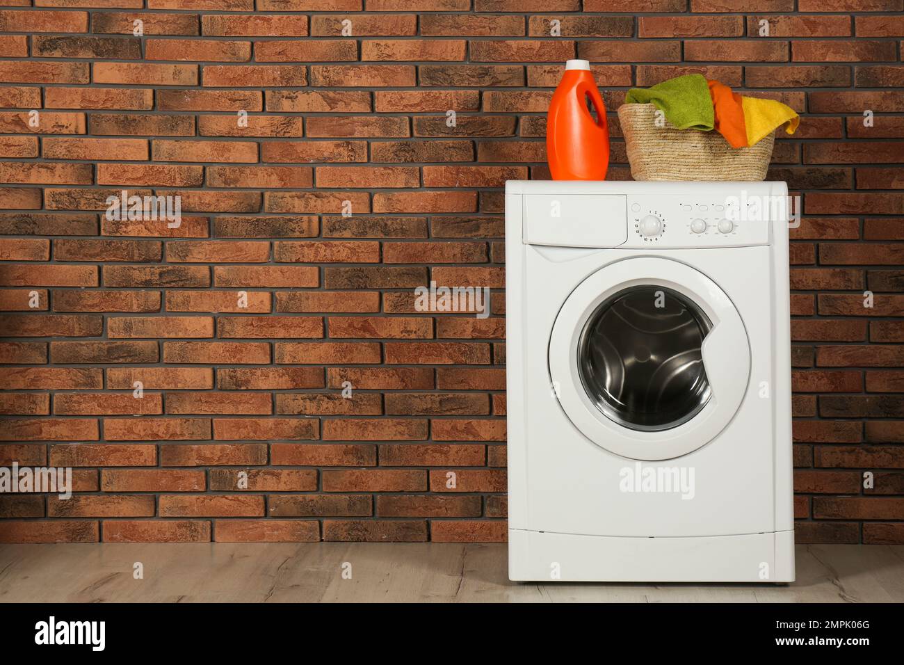 Modern washing machine with detergent and laundry basket near brick ...