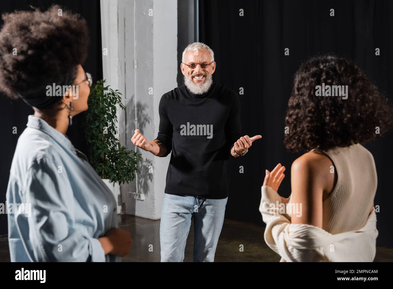 cheerful grey haired man pointing with thumb near multiethnic actresses ...