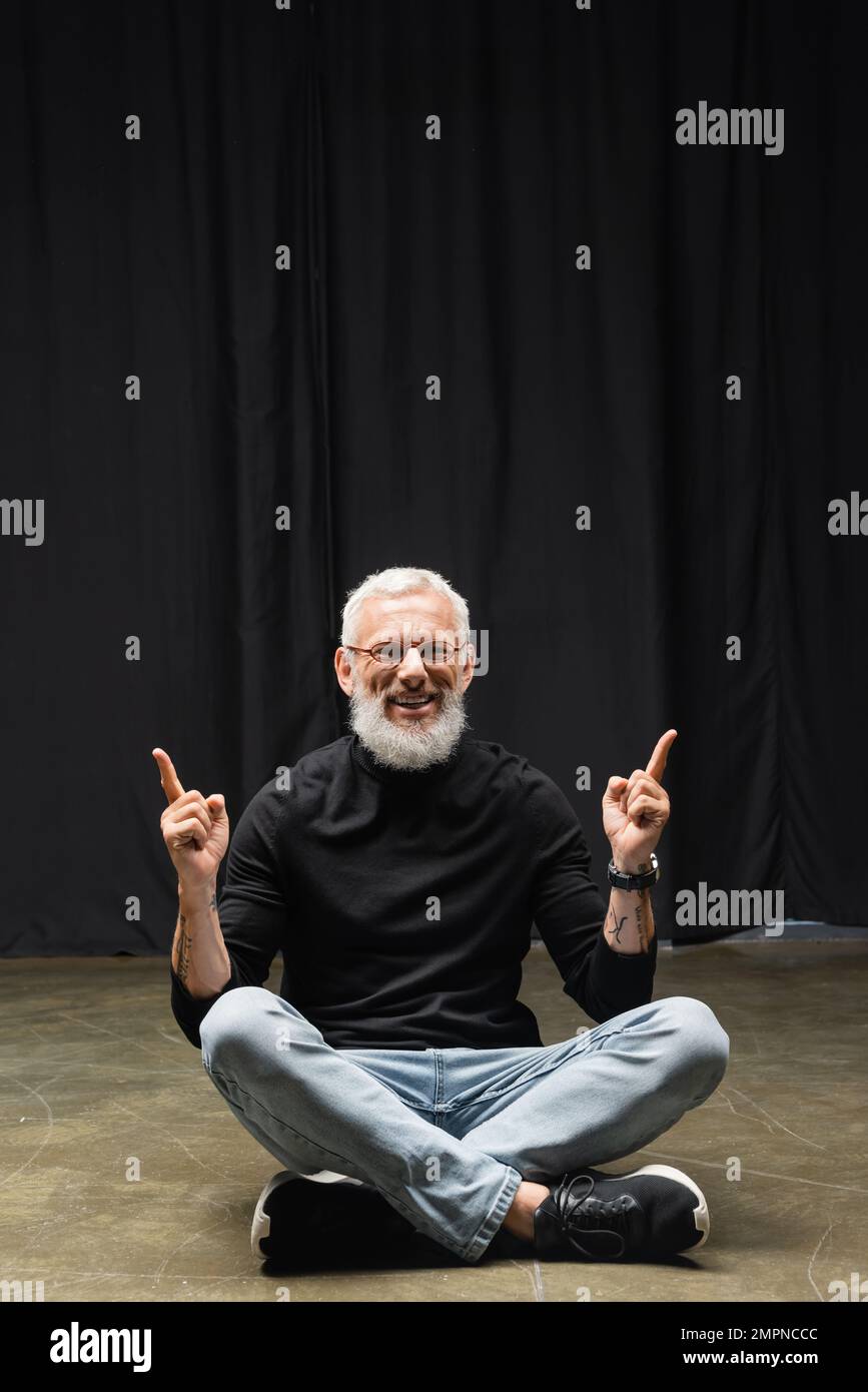 cheerful tattooed actor pointing up with fingers while sitting on ...