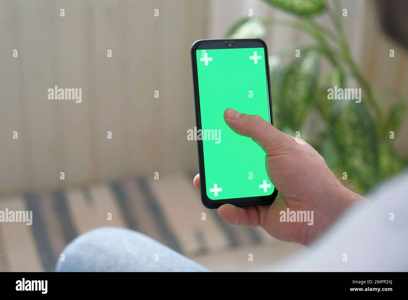 Young man sitting at home holding smartphone green mock-up screen in hand. Male person using ...