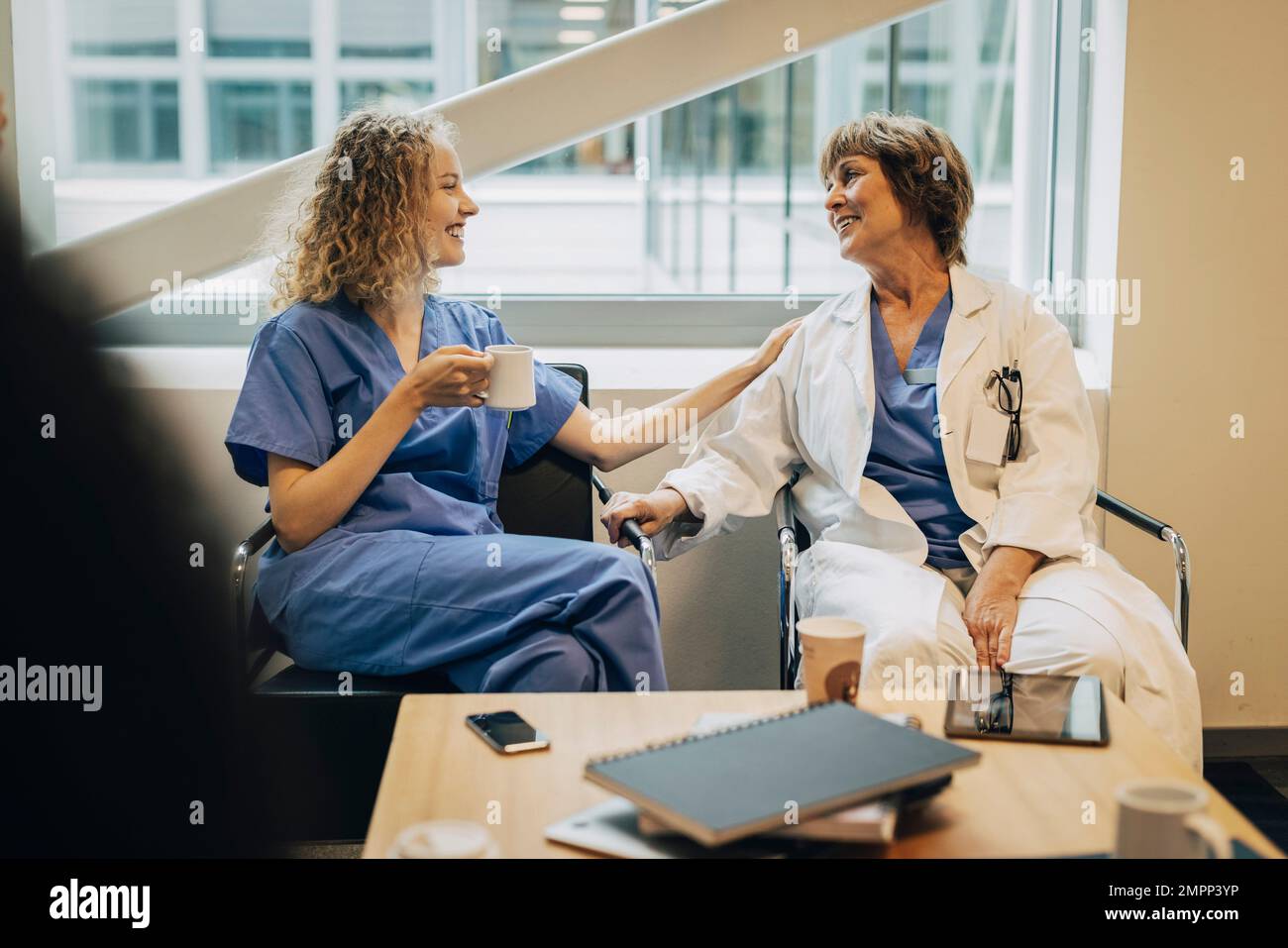 Smiling young nurse talking to mature physician while sitting on chair ...