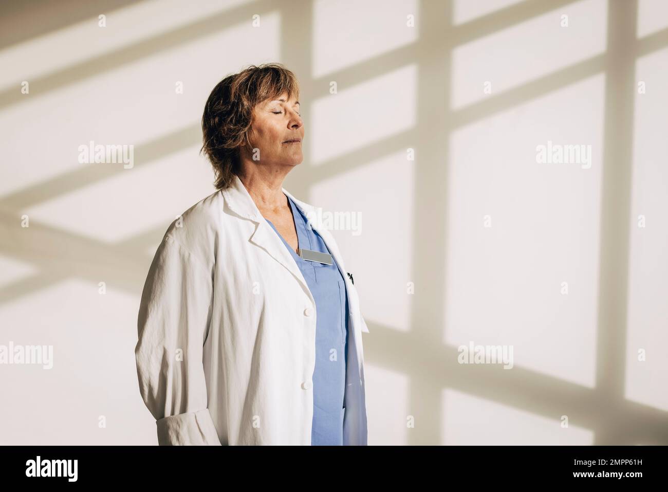 Mature female physician standing with eyes closed against wall with ...