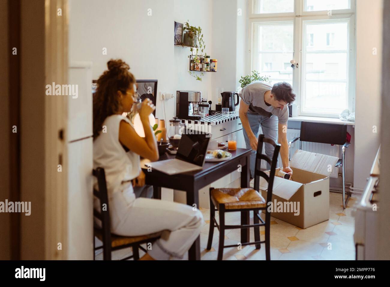 Man unboxing cardboard box in kitchen with girlfriend sitting on dining ...