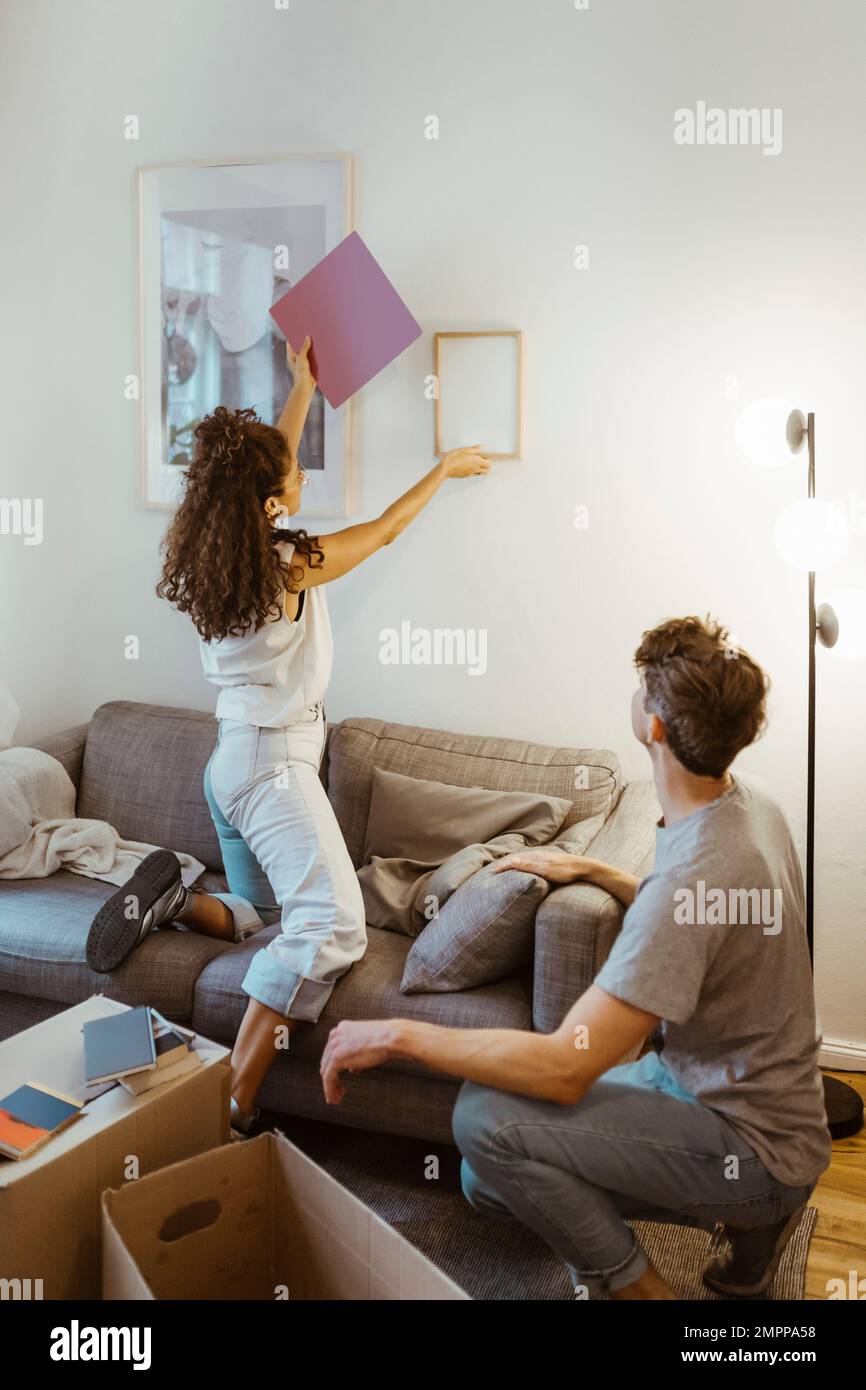 Side view of man looking at girlfriend mounting photo frame on wall ...