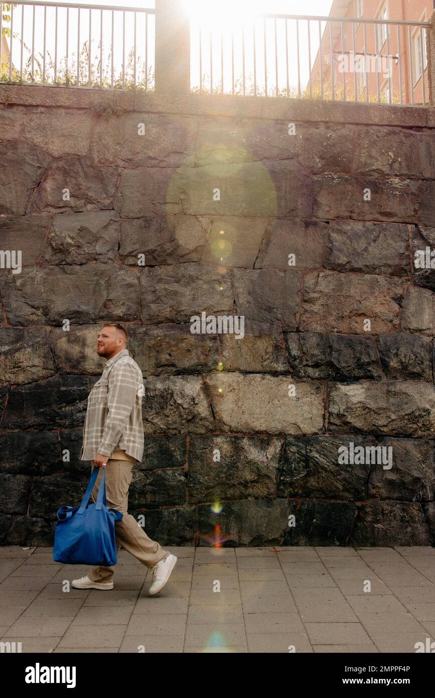 Side view of man walking on footpath in front of wall during sunny day ...
