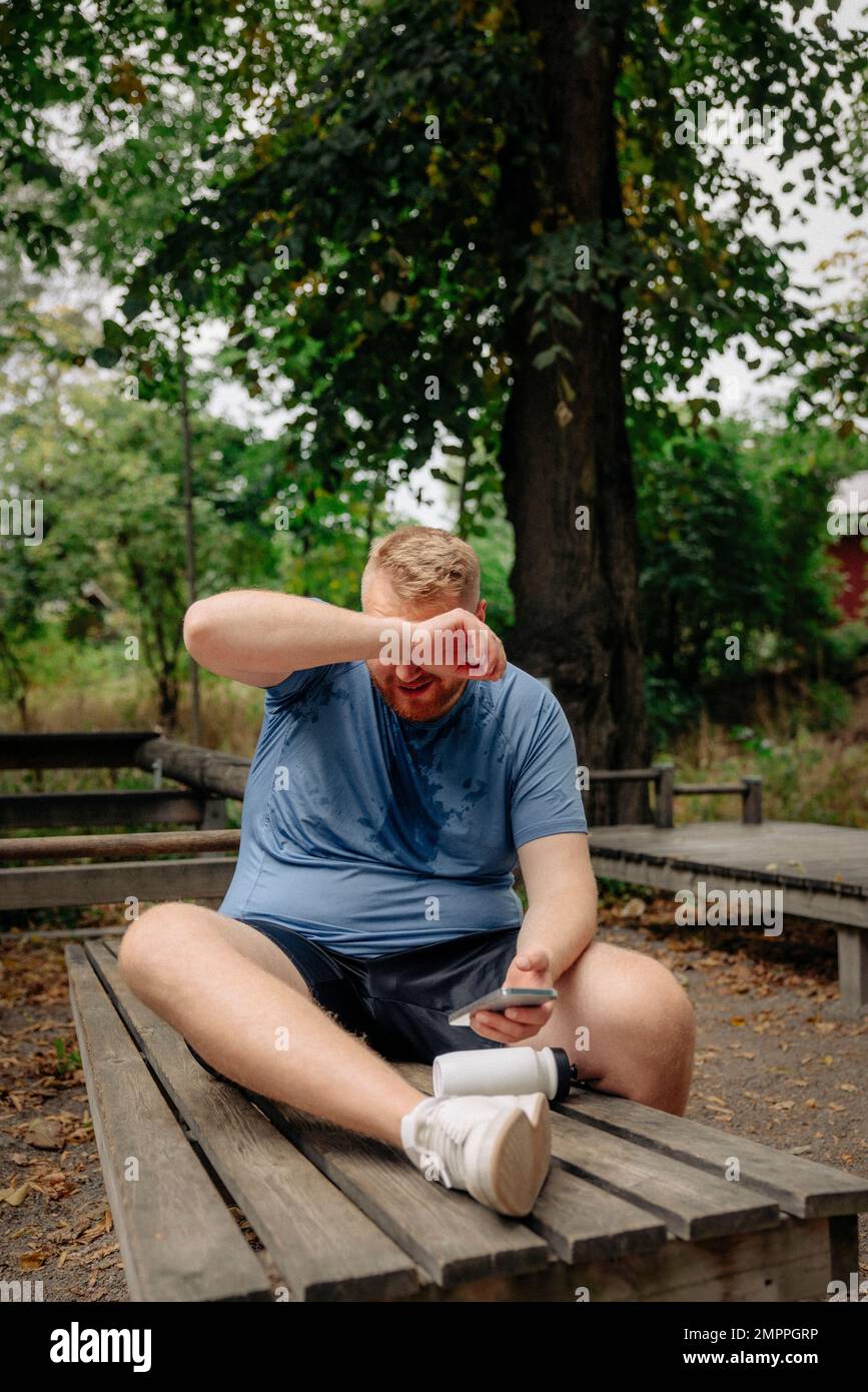 Obese man with smart phone wiping sweat while sitting on bench at park ...