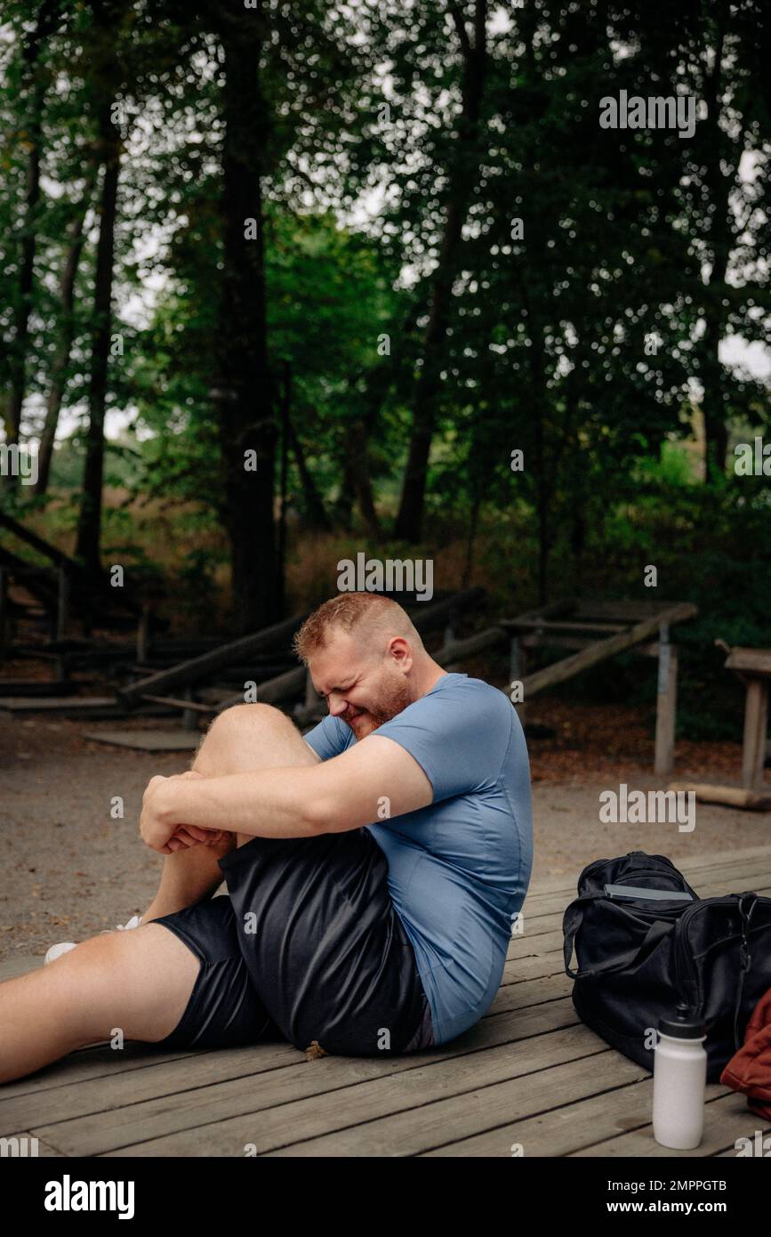 Side view of obese man stretching legs while sitting on bench at park ...