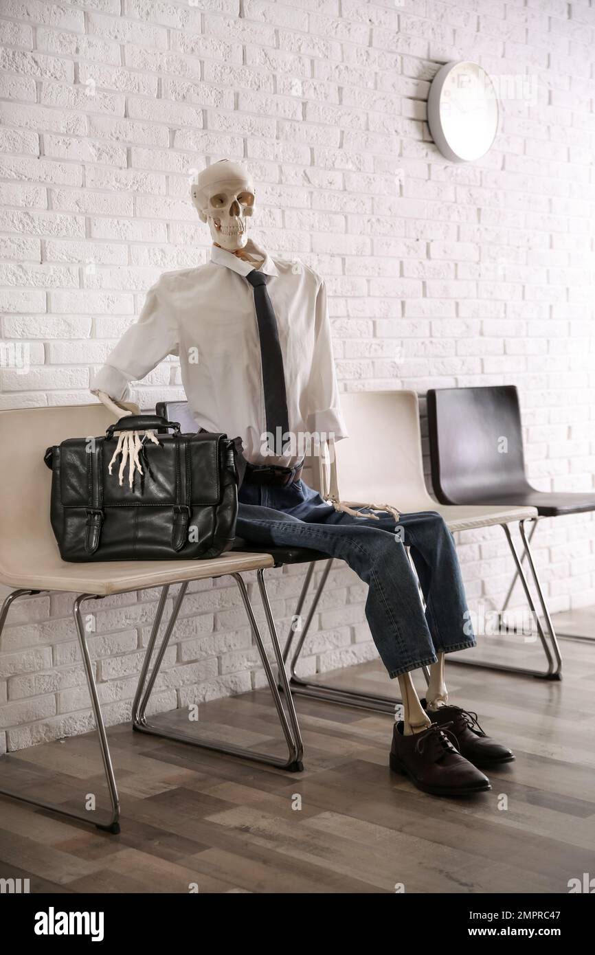 Human skeleton in office wear sitting on chair near brick wall indoors ...