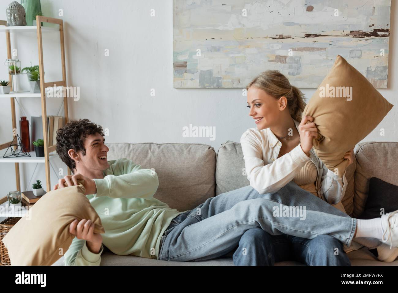 positive young couple smiling while having pillow fight in living room ...