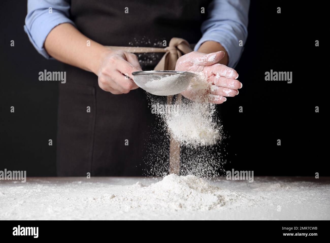 Woman sifting wheat flour at table against black background, closeup ...