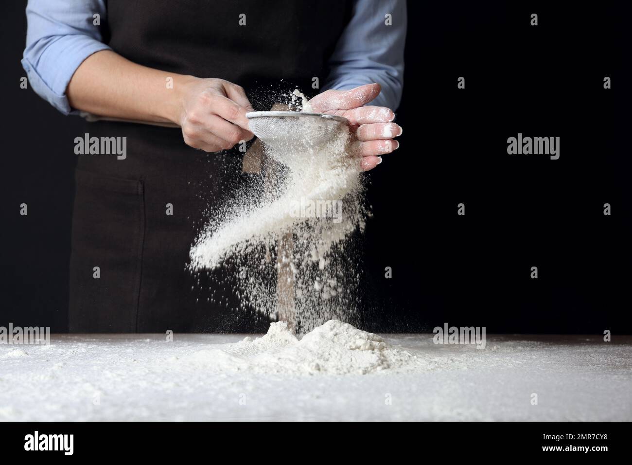 Woman sifting wheat flour at table against black background, closeup ...