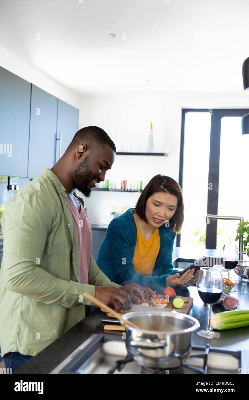 Vertical of happy diverse couple cooking together and using tablet in ...