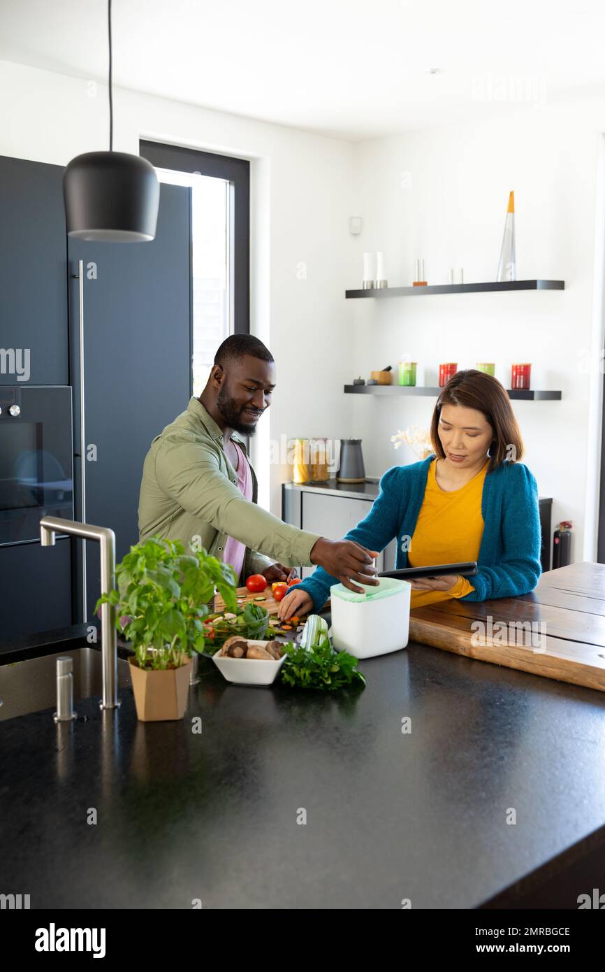 Vertical of happy diverse couple cooking, using tablet and composting ...
