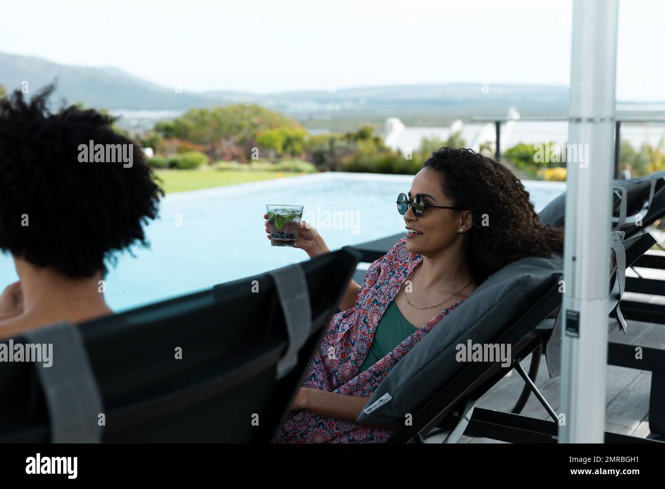 Happy biracial couple relaxing on sun loungers with drinks by pool ...