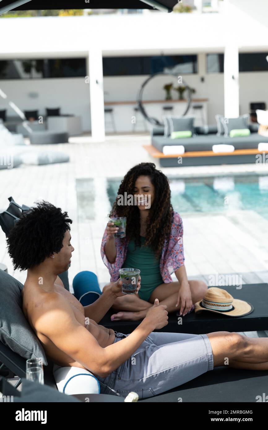 Vertical of happy biracial couple relaxing on sun loungers with drinks ...