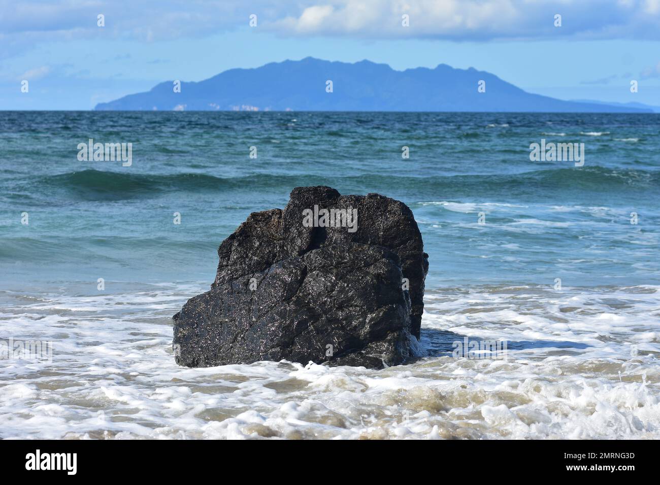 Solitary black volcanic rock washed by oceanic surf with island in ...