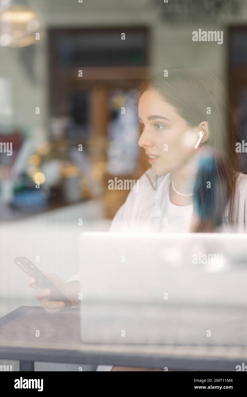 Young woman working with laptop in cafe. View through the window with ...