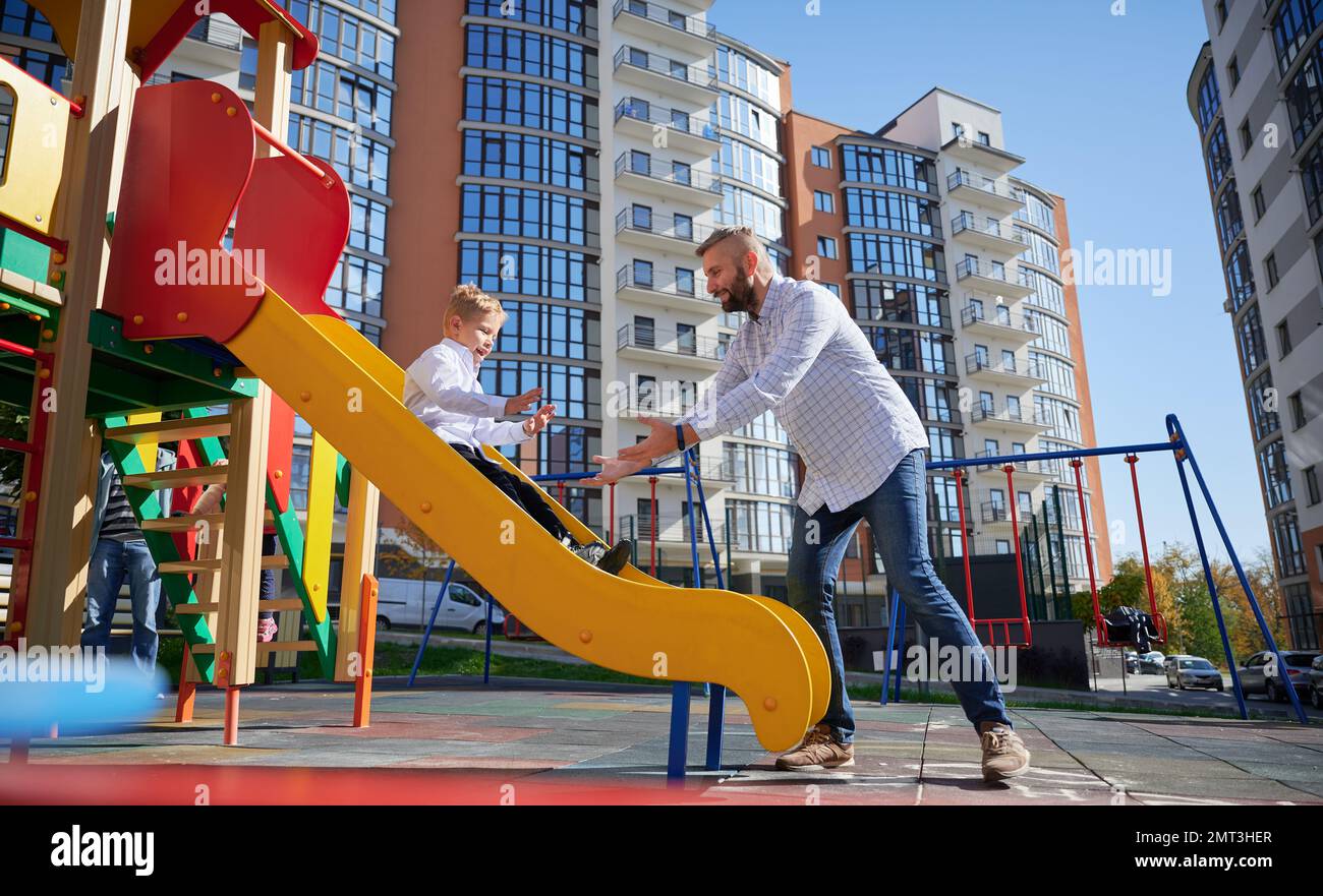 Handsome dad catching male kid riding on slide at sunny day at modern ...