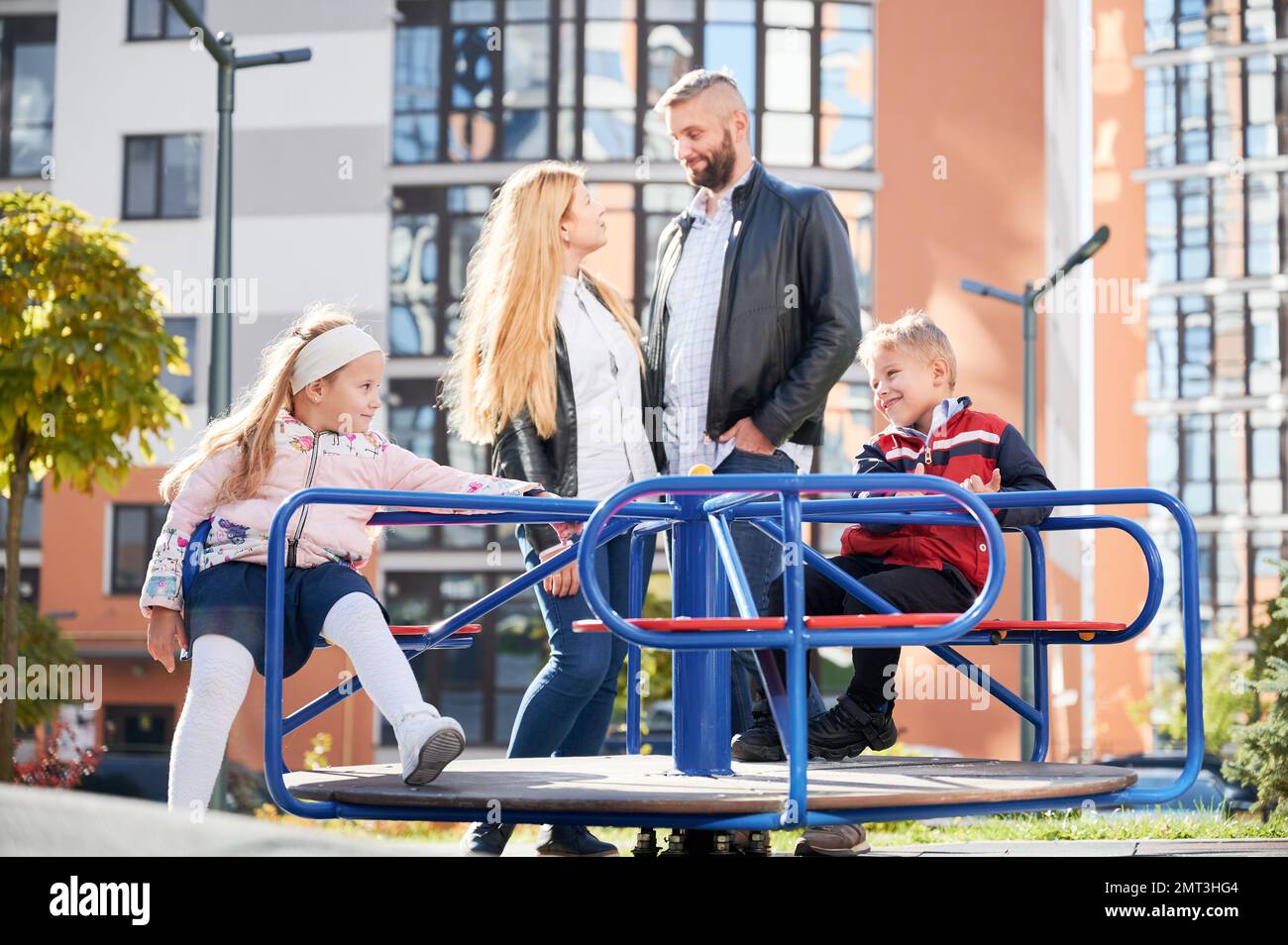 Happy parents hanging out with kids, while children swinging at playground. Front view of two ...