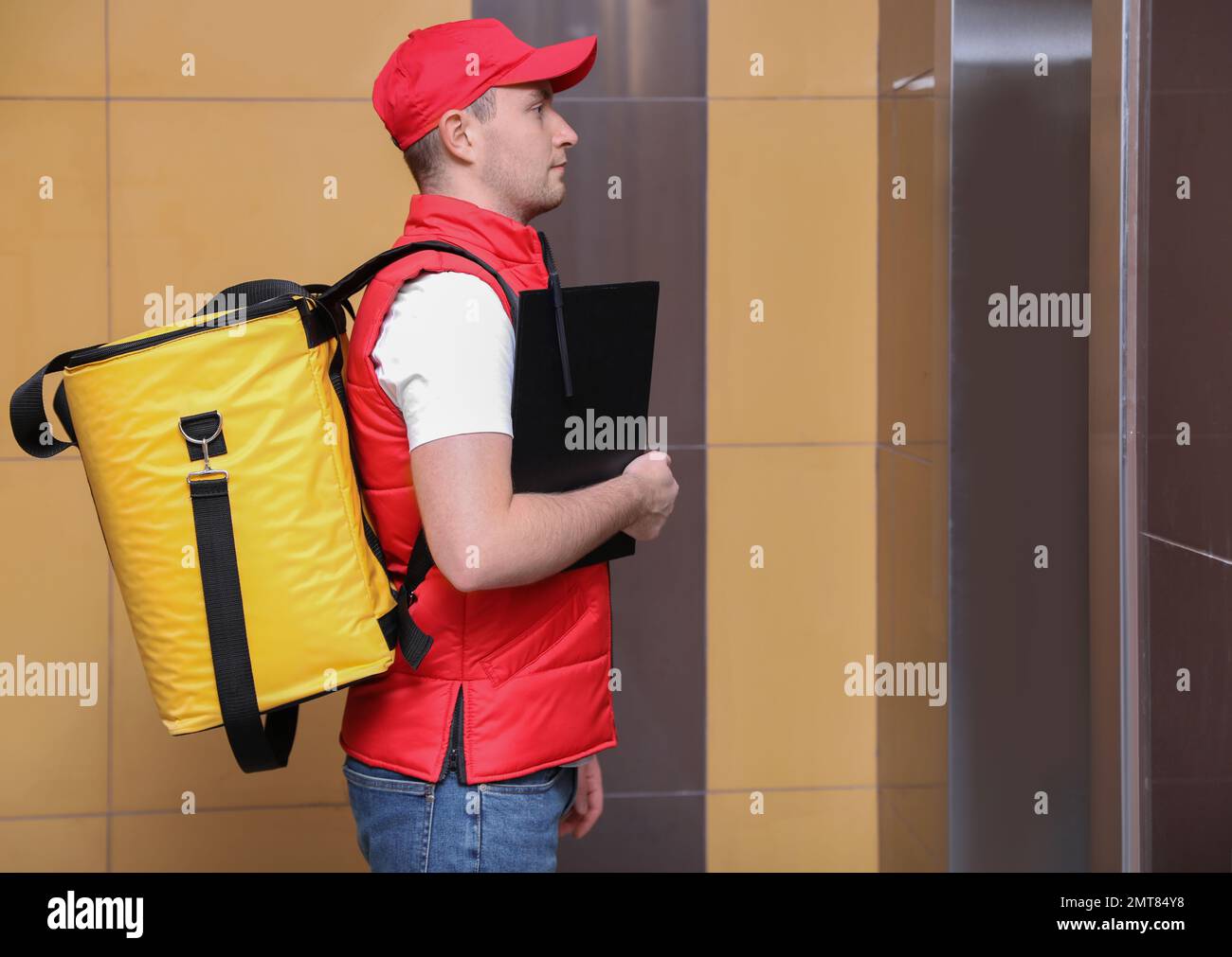 Male courier with thermo bag and clipboard waiting for elevator. Food ...