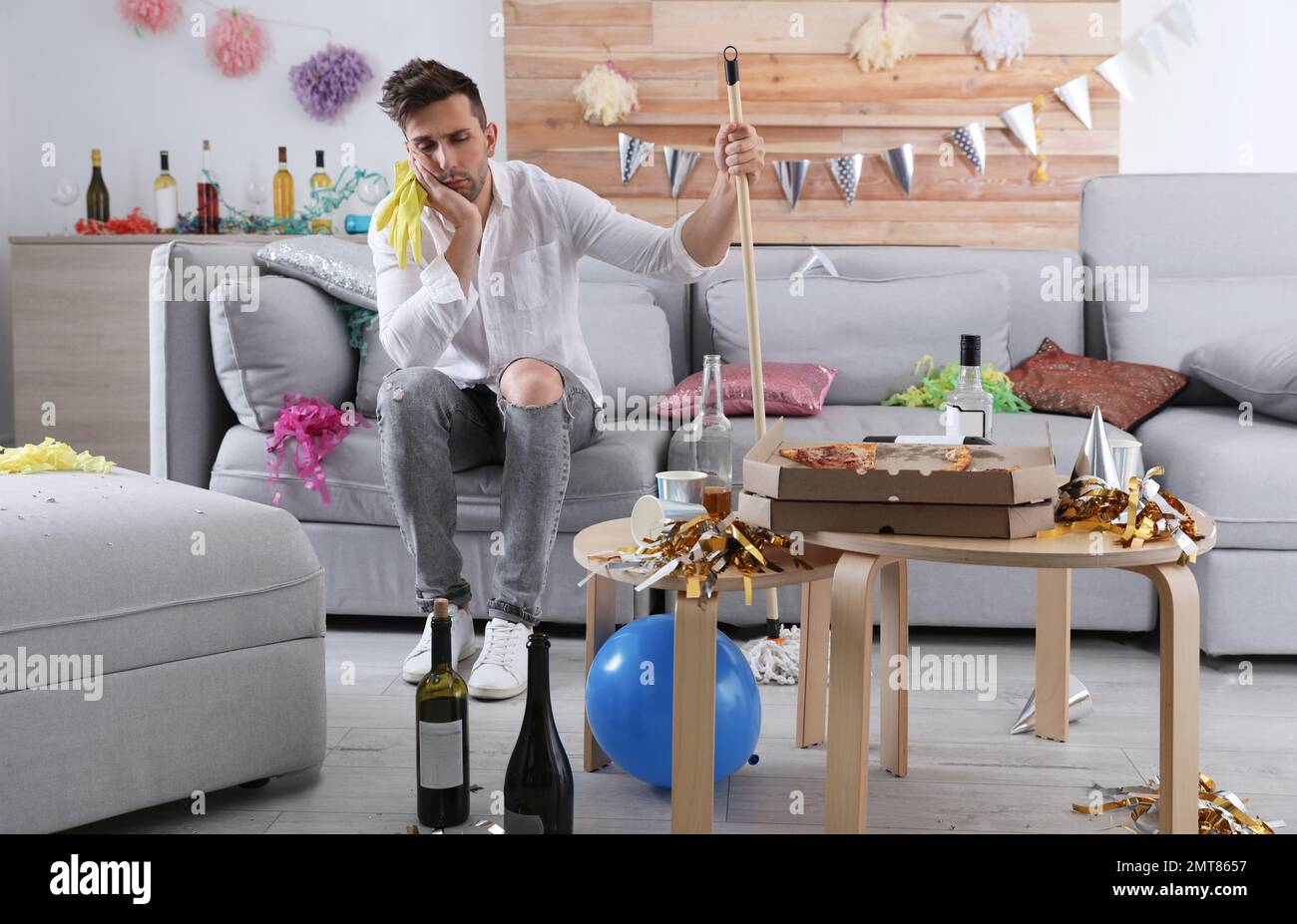 Young man with mop suffering from hangover in messy room after party ...